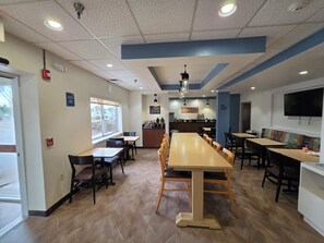 Um restaurante com uma longa mesa de madeira no centro, rodeada por cadeiras. Há várias mesas e cadeiras, um balcão com uma máquina de café e uma televisão na parede.