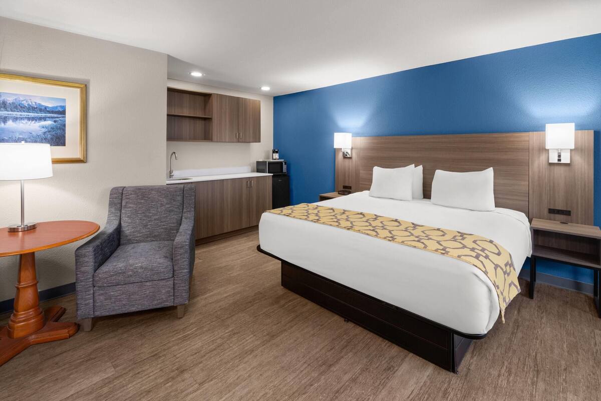 Quarto de hotel com cama, uma cadeira, mesa, micro-ondas, pia e um quadro na parede.