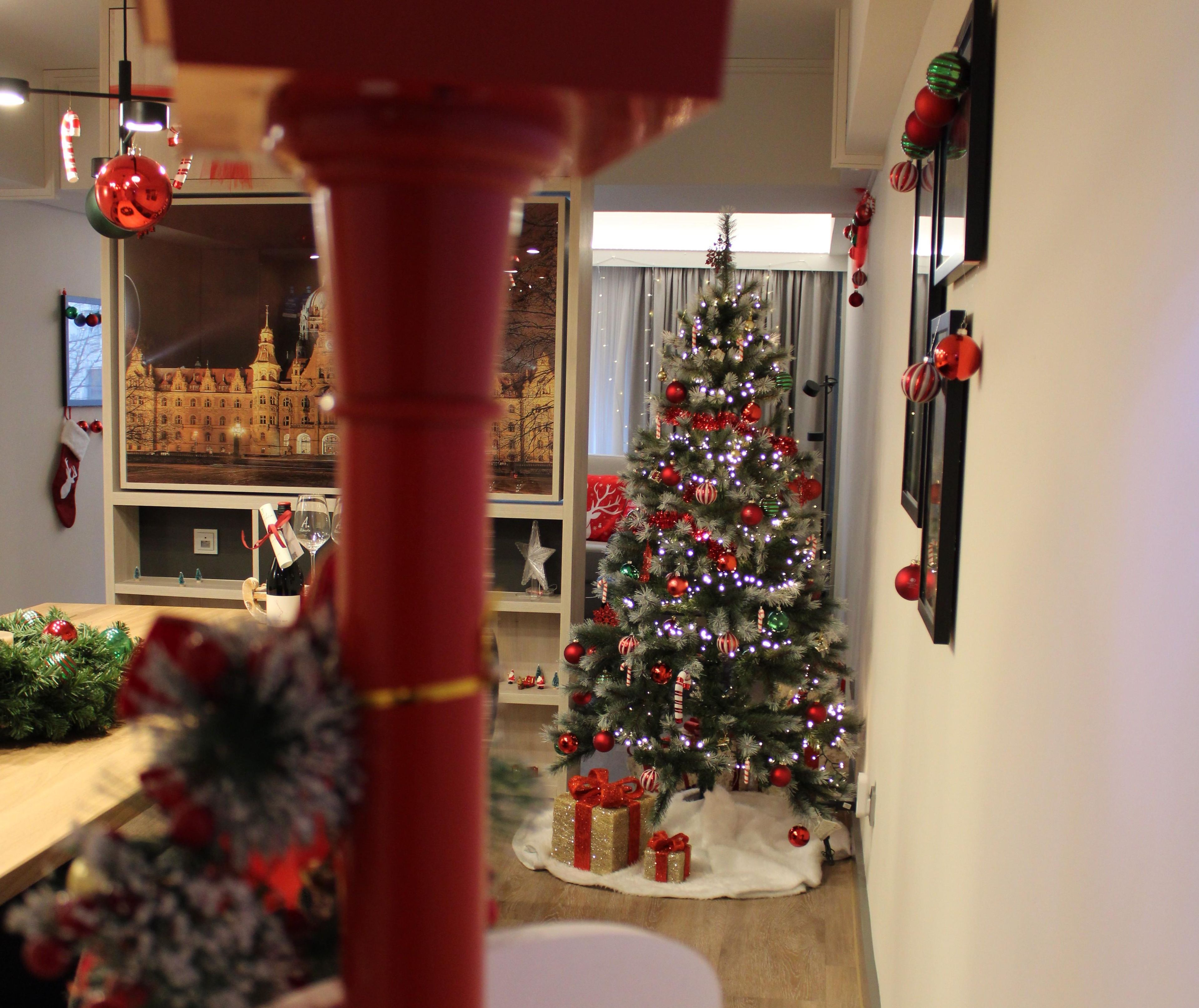 Um quarto com árvore de Natal decorada com enfeites vermelhos e verdes, um quadro pendurado na parede e uma garrafa de champanhe sobre uma mesa.