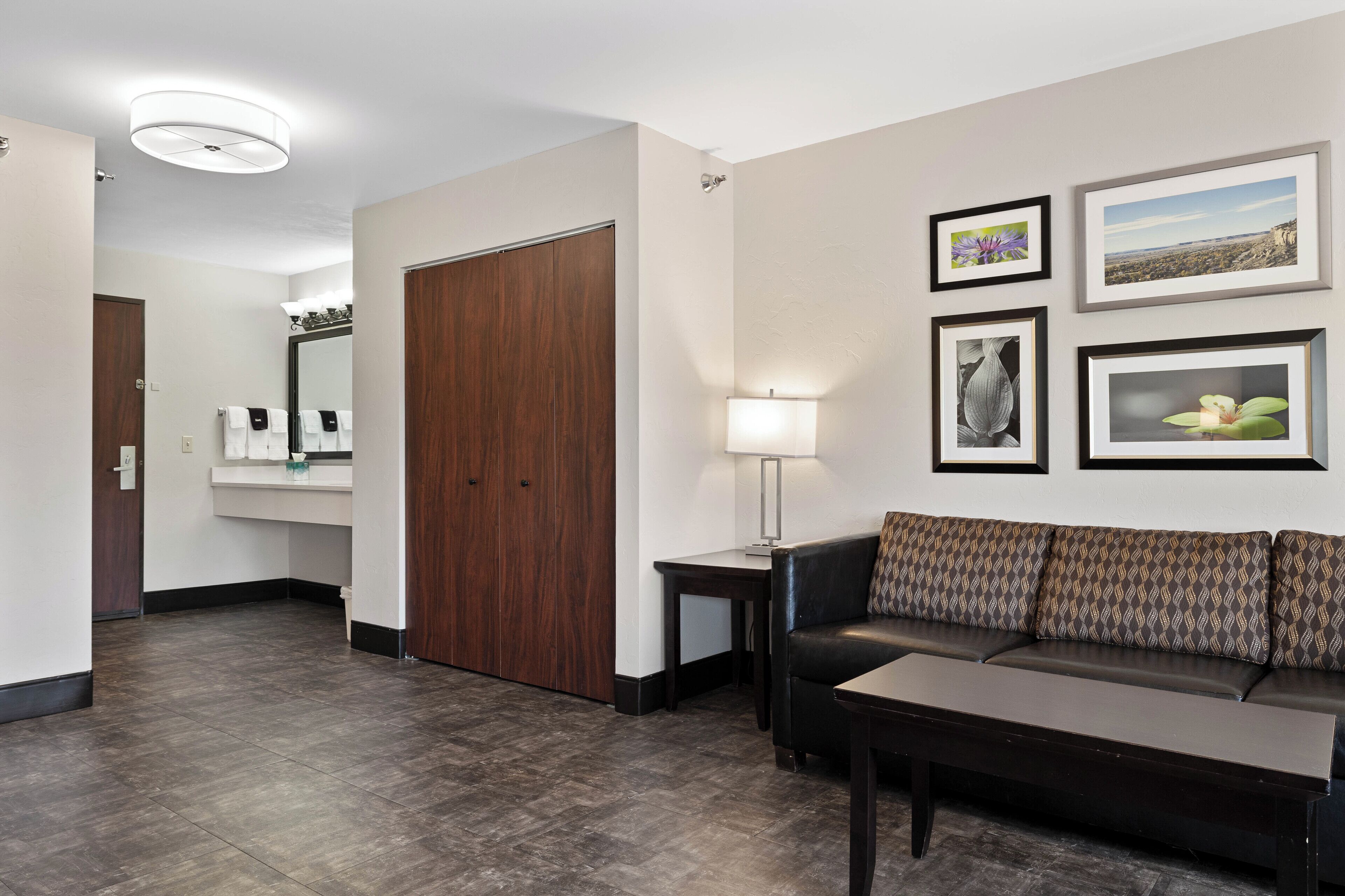 Quarto de hotel com um sofá marrom escuro, uma mesinha, uma luminária, um espelho e um guarda-roupa. Há quadros nas paredes.