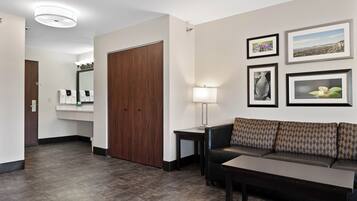Quarto de hotel com um sofá marrom escuro, uma mesinha, uma luminária, um espelho e um guarda-roupa. Há quadros nas paredes.