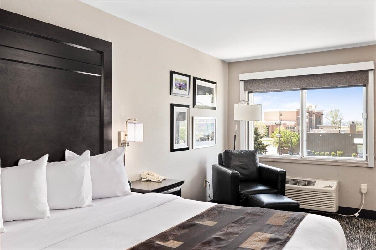 Quarto de hotel com uma cama grande, uma escrivaninha com luminária, uma cadeira e uma janela com vista para a cidade.