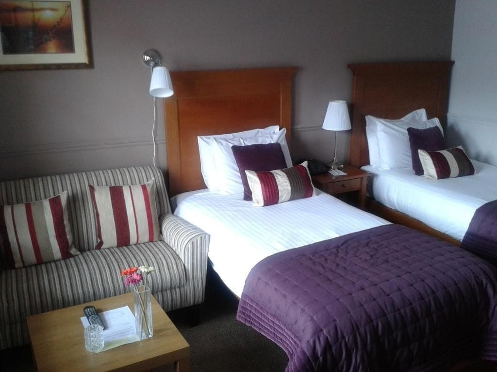 Quarto de hotel com duas camas de solteiro, um sofá, uma mesinha com um vaso de flores e uma luminária.