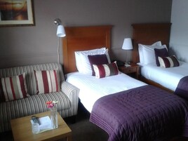 Quarto de hotel com duas camas de solteiro, um sofá, uma mesinha com um vaso de flores e uma luminária.