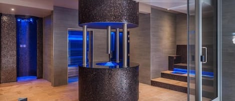 Uma área de spa moderna com um elemento aquático circular em mosaico, um elevador de vidro e uma receção.