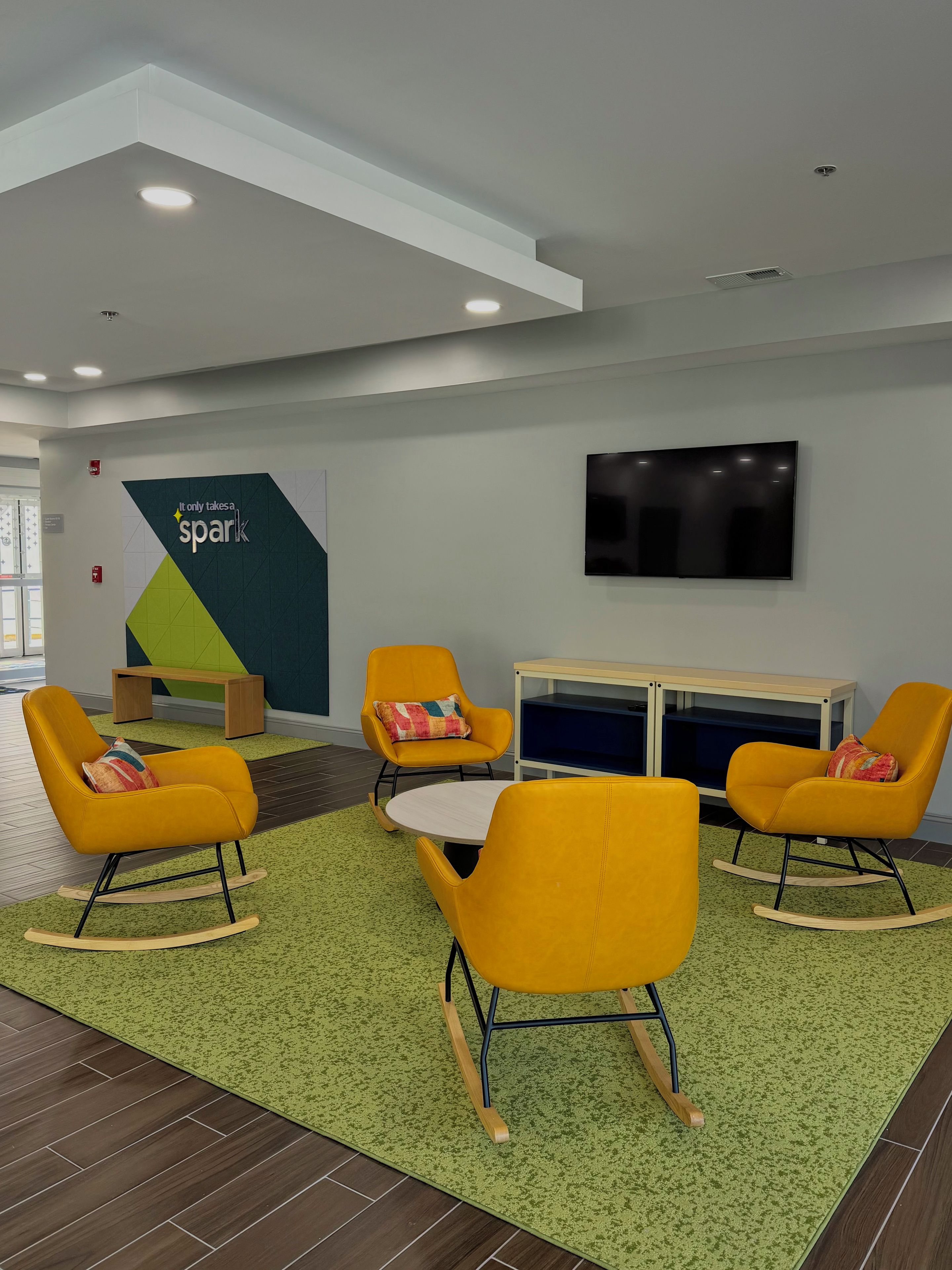 Um interior moderno com área de estar, incluindo cadeiras de balanço amarelas e uma mesa redonda, uma TV de tela plana na parede, e um tapete verde estampado no chão.