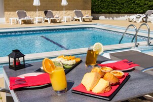 Mesa à beira da piscina com um copo de sumo de laranja, um copo de chá gelado, um prato de pão e um tabuleiro de fruta.