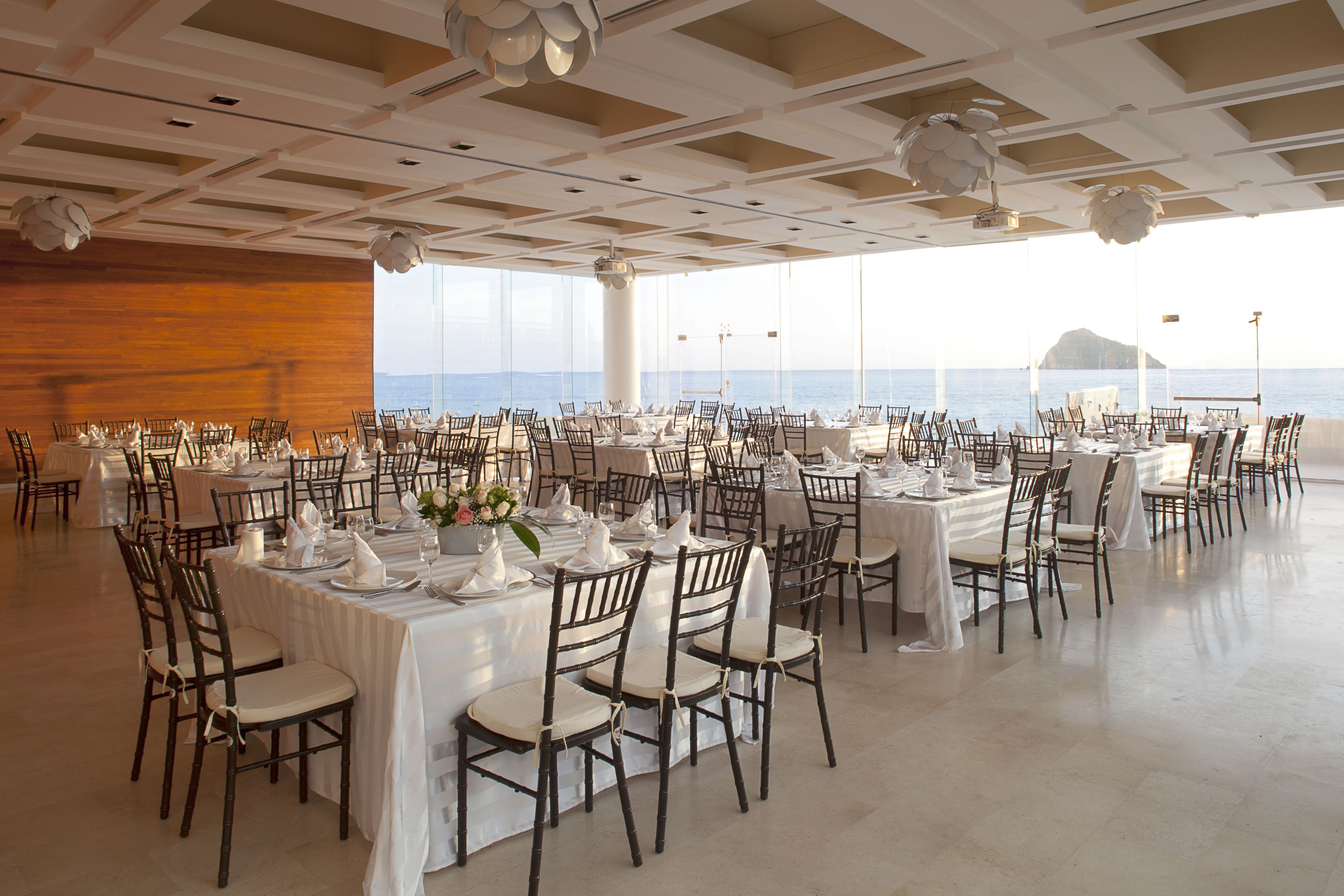 Uma sala de jantar ampla com várias mesas redondas preparadas para um evento formal, grandes janelas com vista para o mar e uma parede de destaque em madeira.