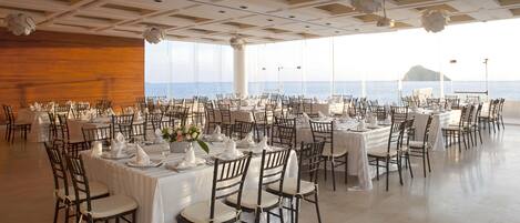 Uma sala de jantar ampla com várias mesas redondas preparadas para um evento formal, grandes janelas com vista para o mar e uma parede de destaque em madeira.