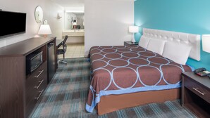 Quarto de hotel com uma cama, escrivaninha com televisão, uma cadeira, luminária de mesa e banheiro visível ao fundo.