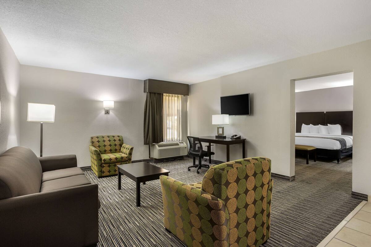 Quarto de hotel com sofá, duas poltronas, escrivaninha com abajur, televisão de tela plana e cama.