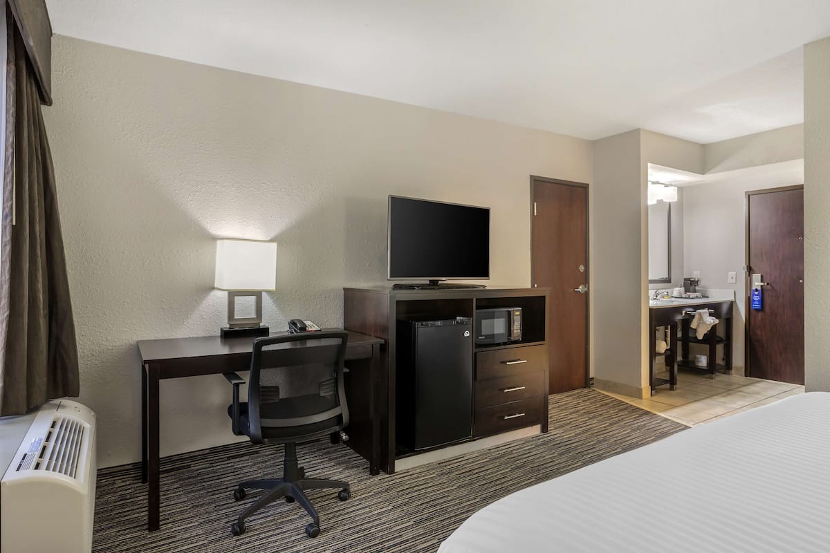 Quarto de hotel com cama, escrivaninha com computador, televisão, micro-ondas, pequeno espaço de cozinha e banheiro.