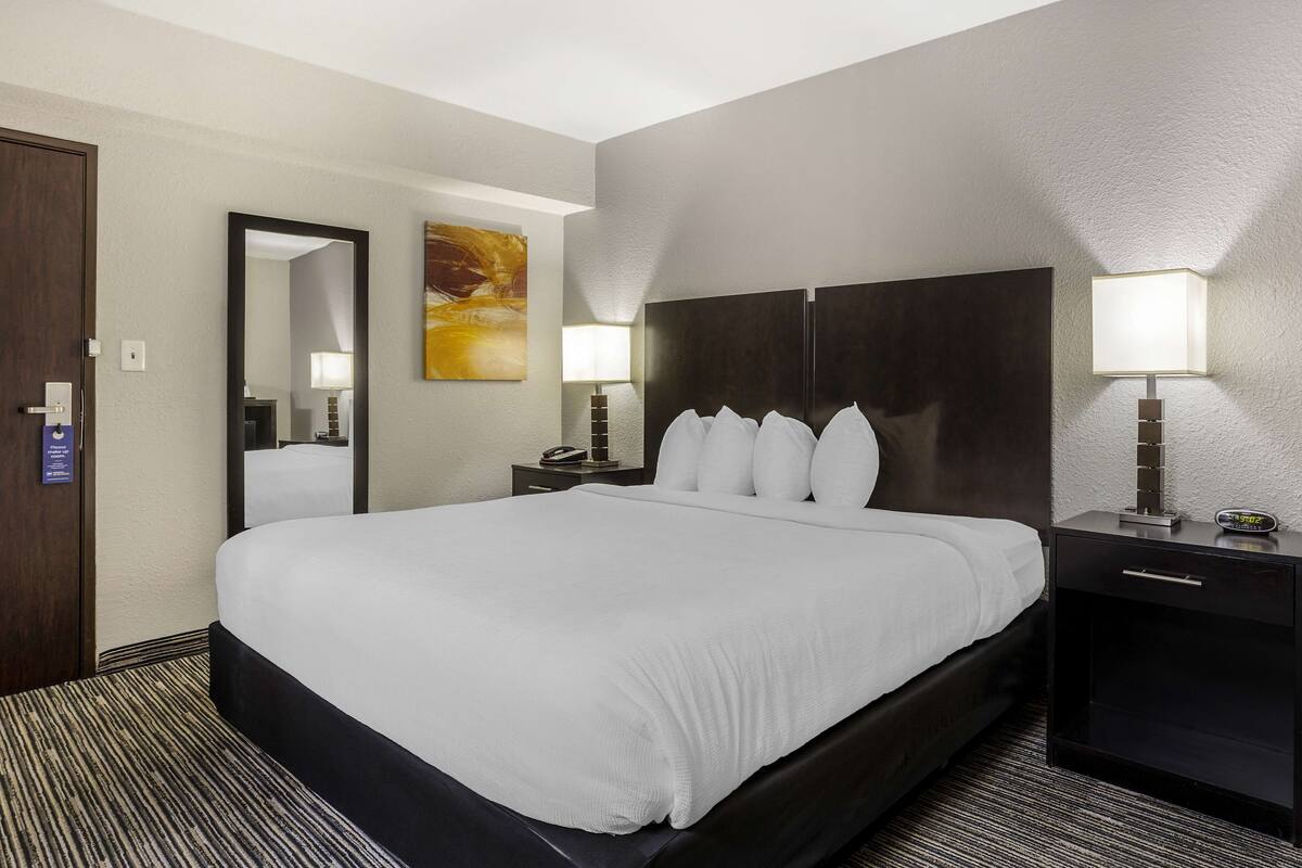 Quarto de hotel com uma cama grande, duas mesas de cabeceira, um criado-mudo, um espelho e uma obra de arte pendurada na parede.
