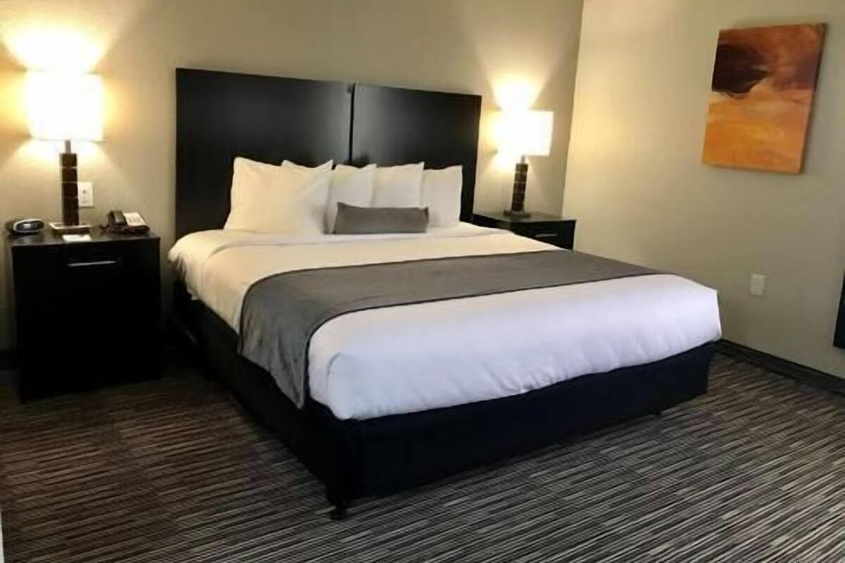 Quarto de hotel com uma cama grande, duas mesas de cabeceira com luminárias, uma mesinha de apoio e um quadro na parede.