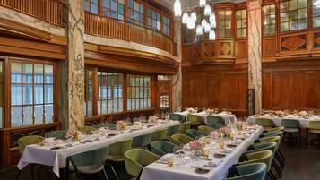 Uma sala de banquetes com longas mesas preparadas para refeições, painéis de madeira e grandes janelas.