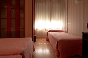 Quarto de hotel com duas camas, uma janela grande com cortinas, um guarda-roupa de madeira e um alto-falante.