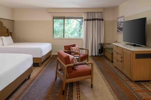 Quarto de hotel com cama, uma cadeira, escrivaninha e televisão, além de uma janela com cortinas.