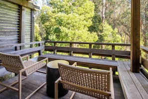 Um deck de madeira com uma mesa e duas cadeiras de vime, com vista para uma área arborizada.
