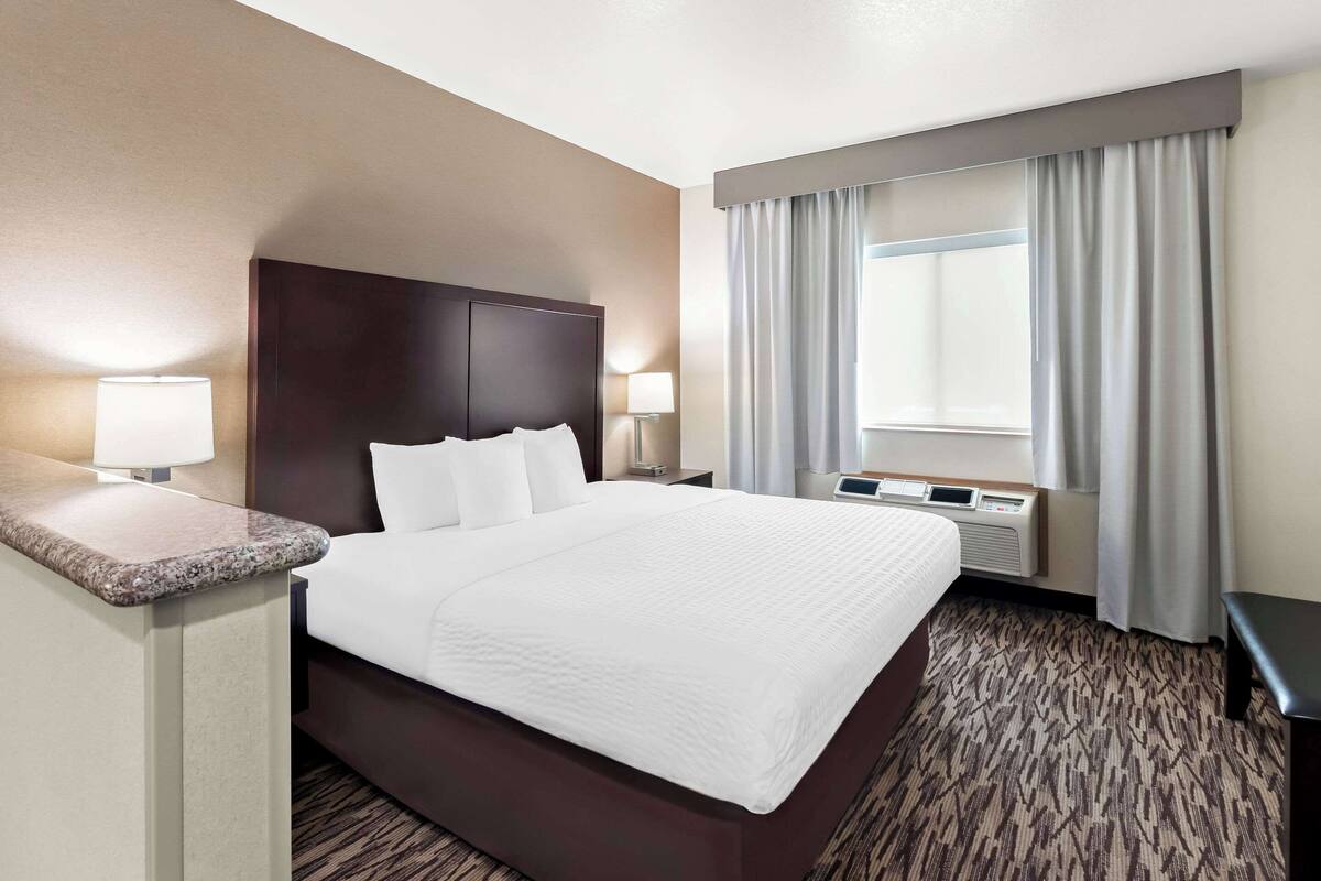 Quarto de hotel com uma cama grande, duas mesas de cabeceira com luminárias, uma janela com cortinas e um ar condicionado fixado na parede.