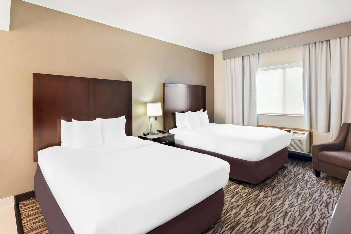 Quarto de hotel com duas camas, uma escrivaninha, uma cadeira e uma janela grande com cortinas.