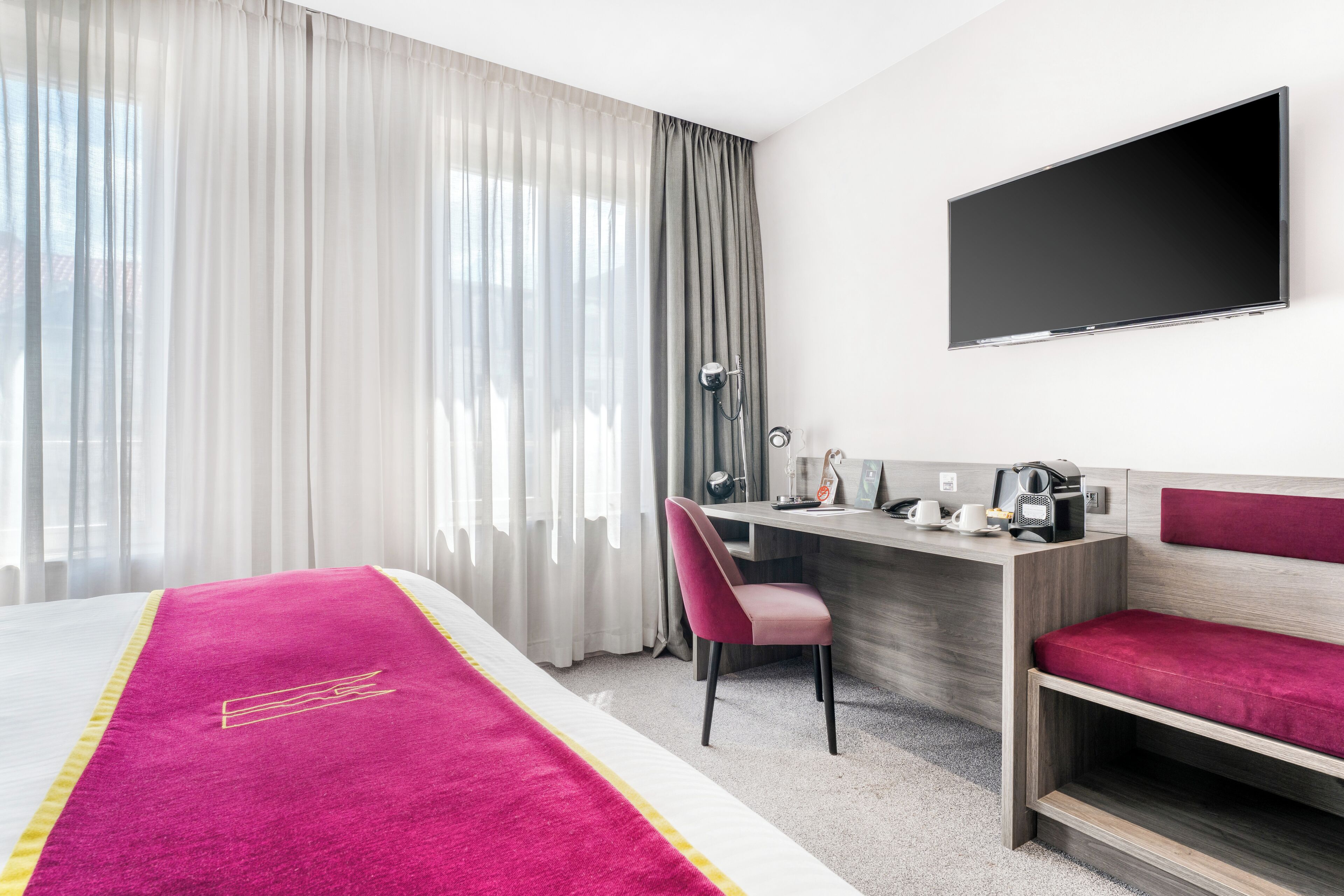 Quarto de hotel com cama, escrivaninha, cadeira, televisão e janela com cortinas.