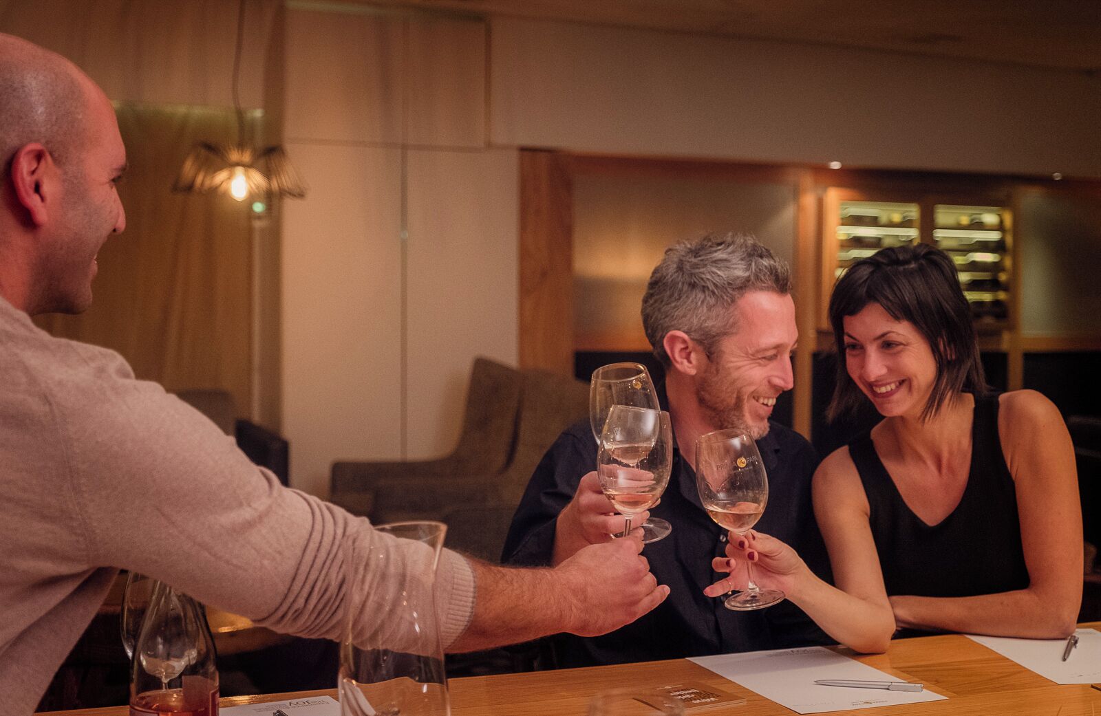 Um grupo de pessoas sentadas à mesa com taças de vinho, em um espaço com mesa e cadeiras de madeira.
