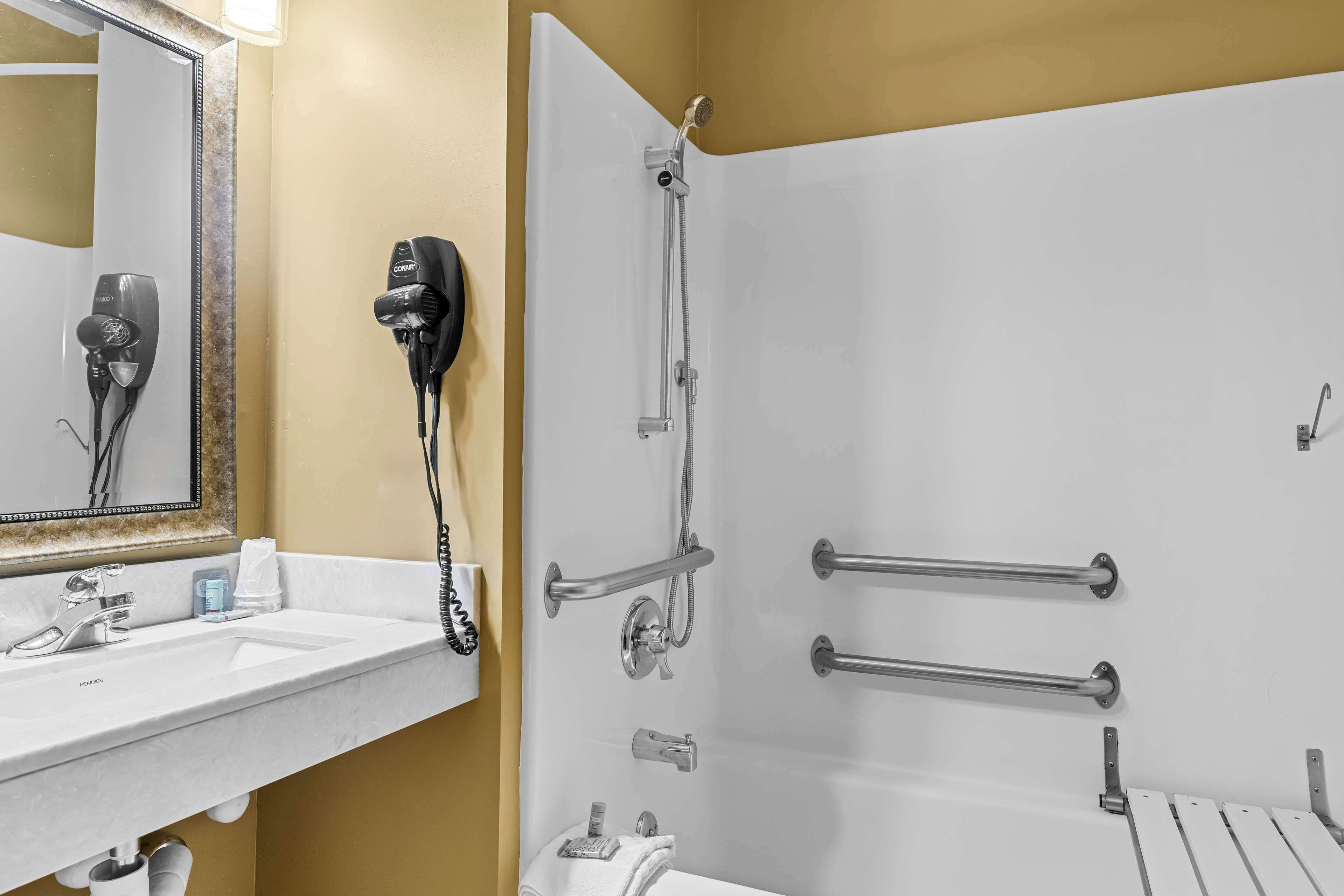 Uma casa de banho com lavatório branco, espelho e secador de cabelo, chuveiro com barras de apoio e um porta-toalhas.