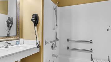 Uma casa de banho com lavatório branco, espelho e secador de cabelo, chuveiro com barras de apoio e um porta-toalhas.