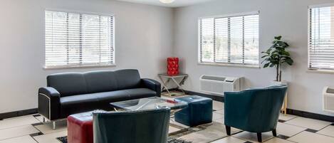 Uma sala de estar moderna com um sofá preto, poltronas azuis e um banquinho vermelho. Há duas janelas com persianas, um radiador e uma planta em vaso.