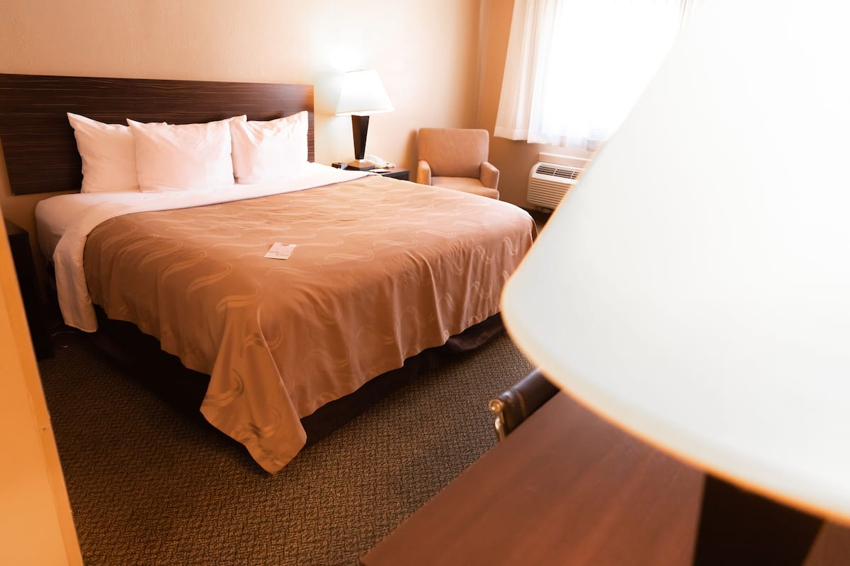 Quarto de hotel com uma cama grande, mesa de cabeceira com luminária, uma cadeira e um aparelho de ar condicionado.