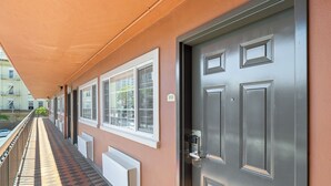 Um corredor com piso xadrez, uma porta de metal com placa de número e janelas com persianas.
