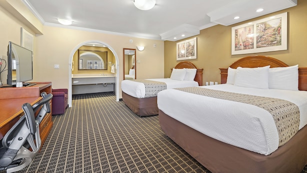 Quarto de hotel com duas camas, uma escrivaninha com computador, um espelho e um banheiro visível através de um arco.