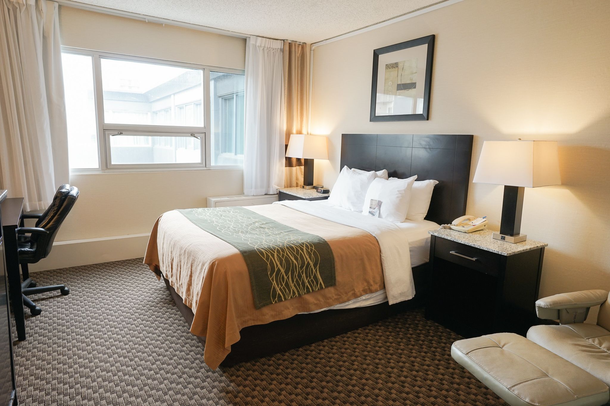 Quarto de hotel com cama, duas mesas de cabeceira com luminárias, escrivaninha com cadeira e janela com cortinas.