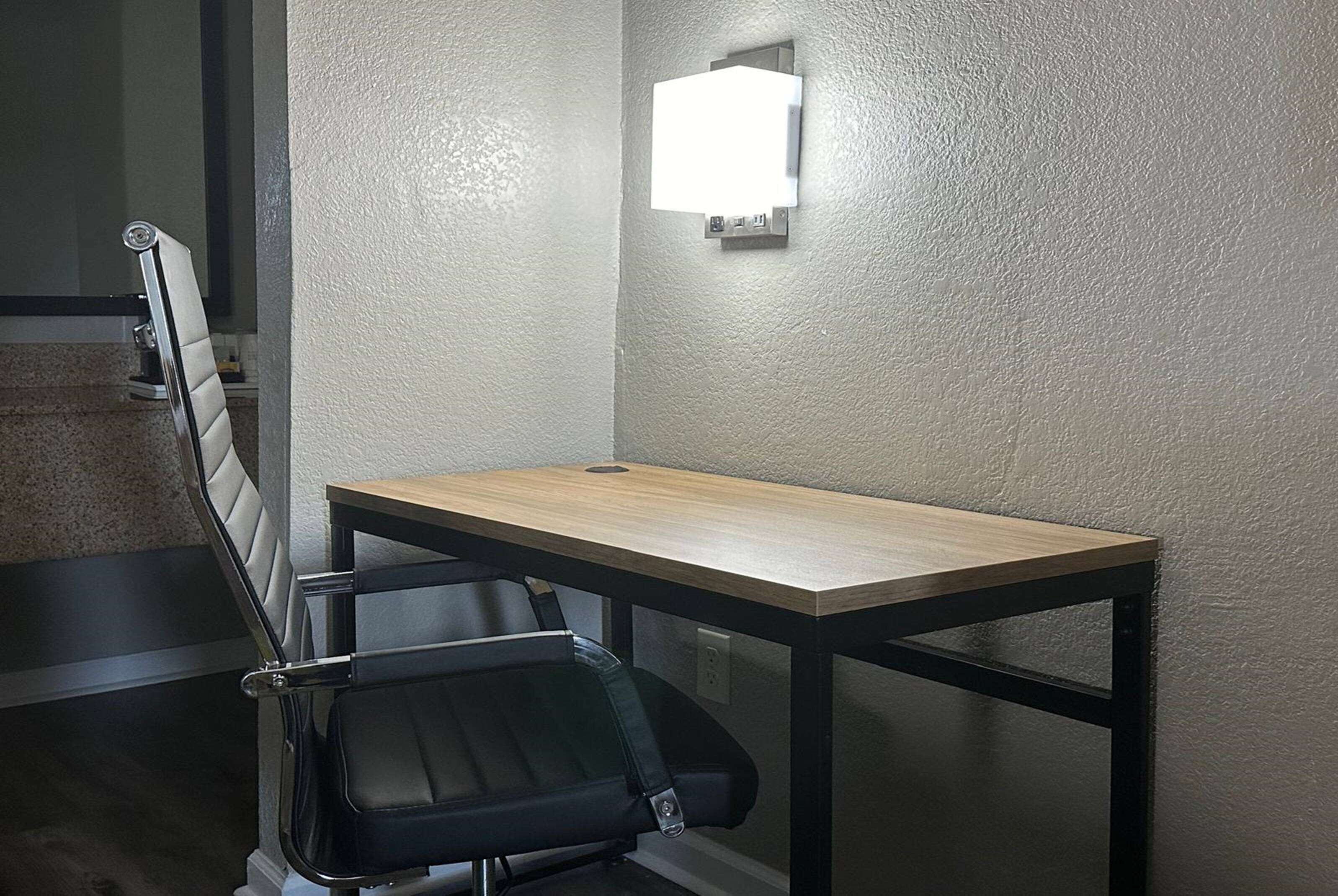 Um quarto pequeno e moderno com uma escrivaninha de madeira e uma cadeira de escritório preta. Há um candeeiro fixado na parede acima da escrivaninha.