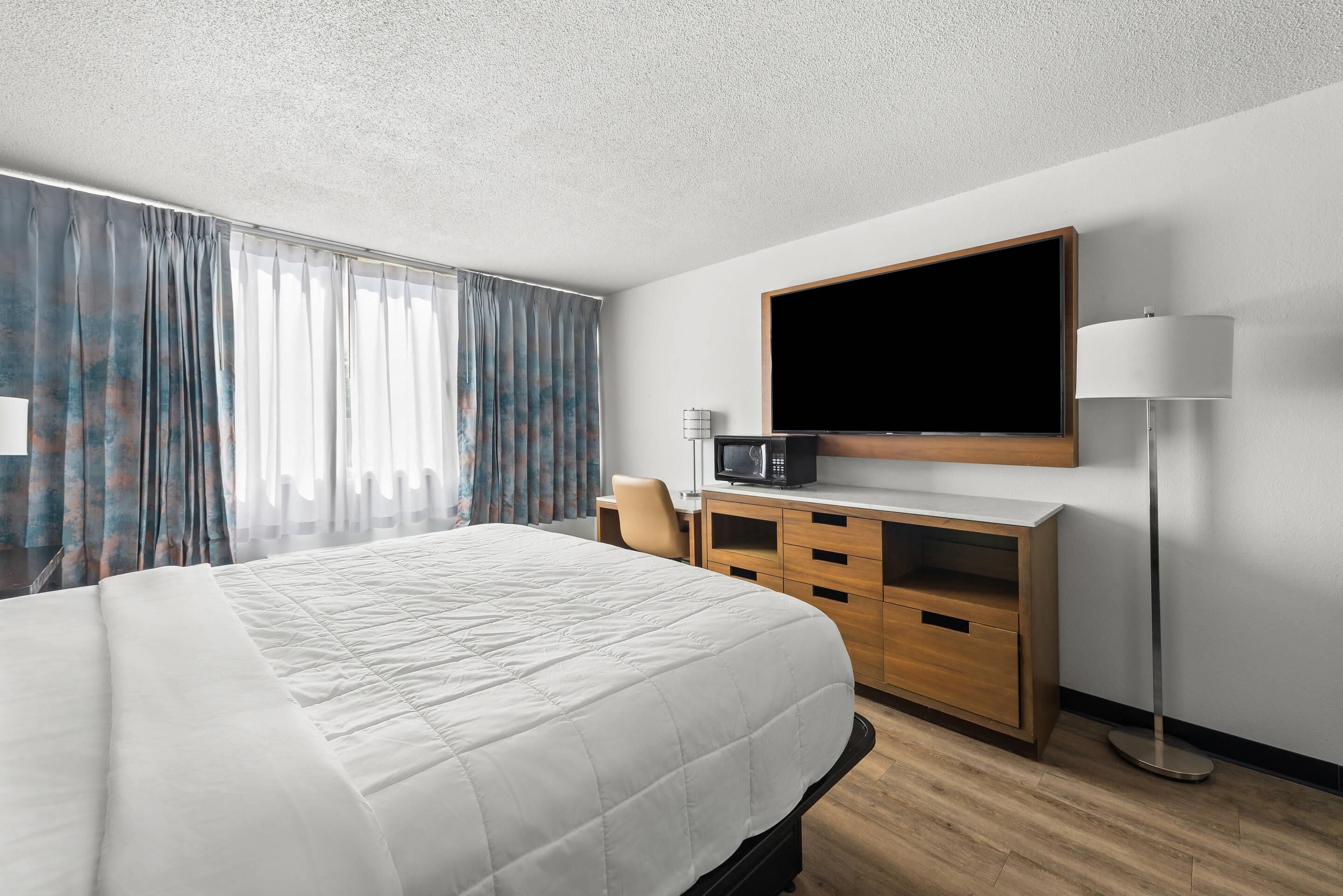 Quarto de hotel com uma cama grande, televisão de tela plana, mesa com cadeira e luminária.
