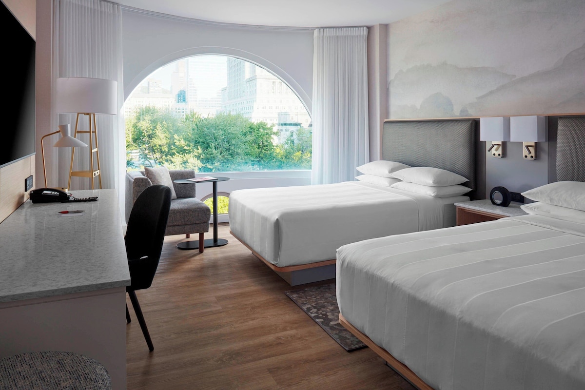 Quarto de hotel com duas camas, uma escrivaninha, uma cadeira, uma mesinha e uma janela grande com vista para a cidade.