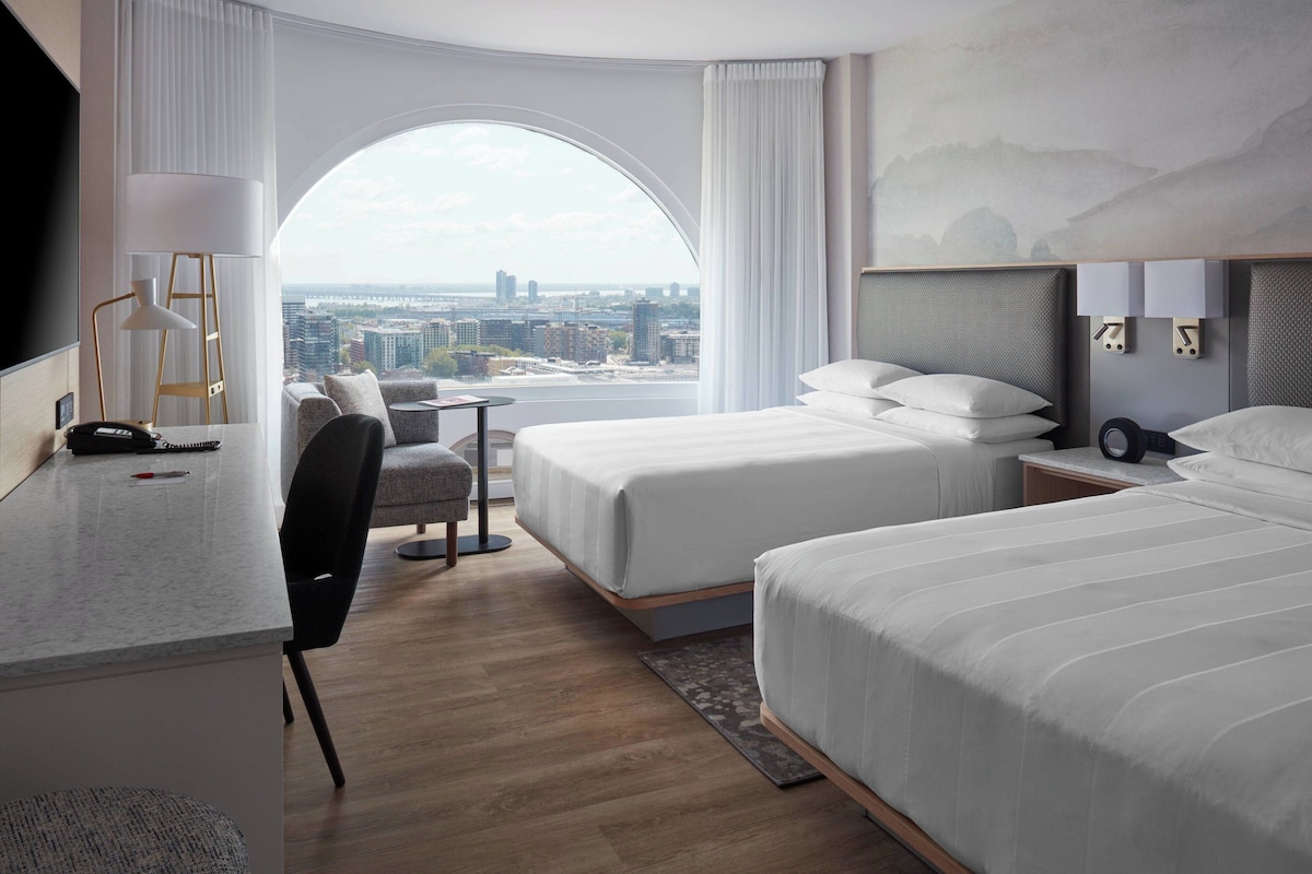 Quarto de hotel com duas camas, uma escrivaninha, uma cadeira, um abajur e vista para a cidade.