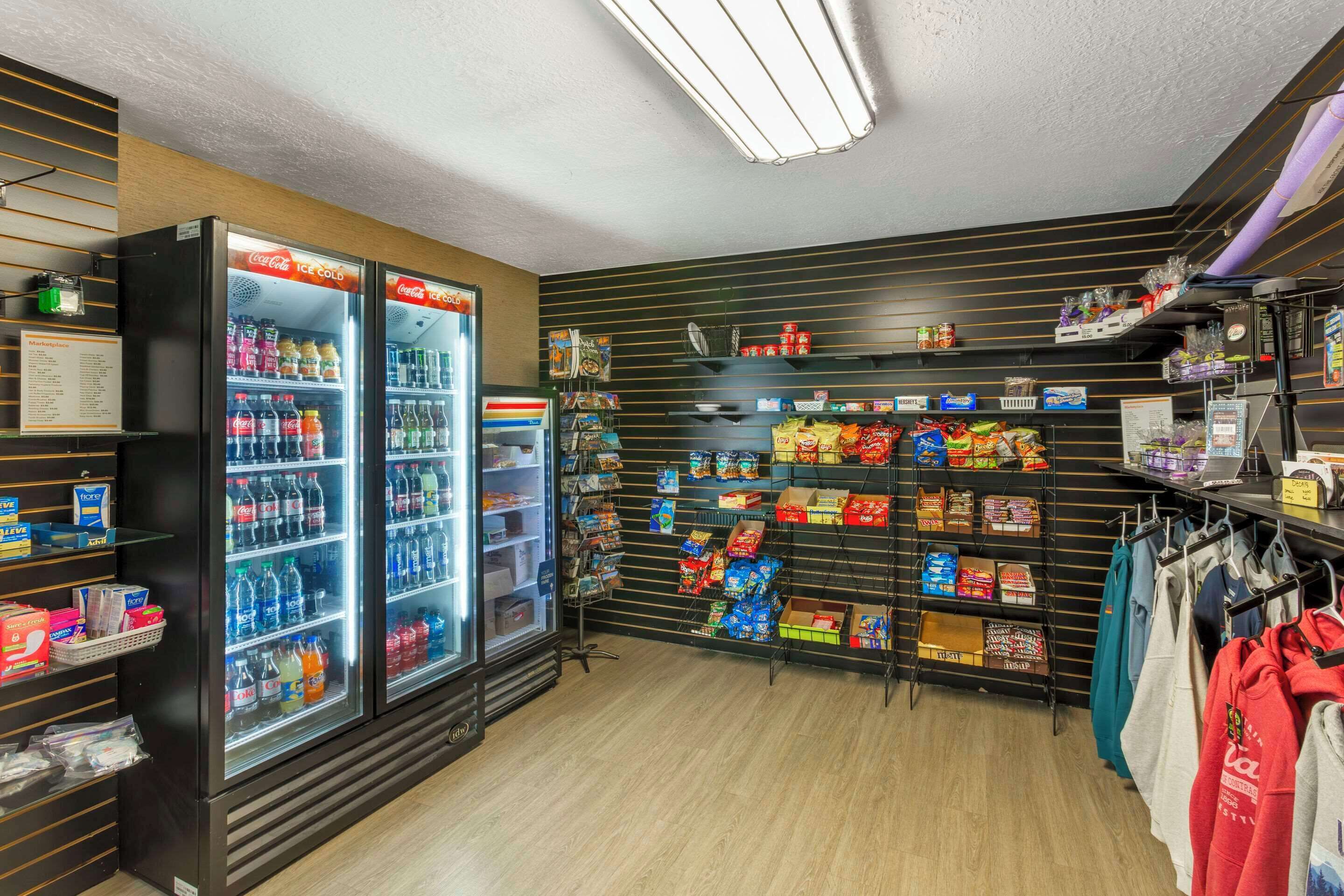 Interior de loja com máquinas de bebidas refrigeradas, prateleiras cheias de lanches e uma arara com roupas.