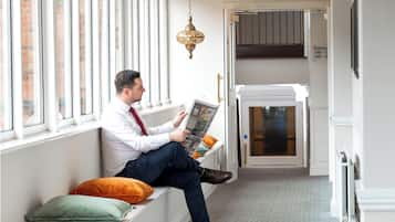 Um homem sentado num banco, lendo uma revista em um corredor com janelas amplas, um radiador e um candeeiro suspenso.