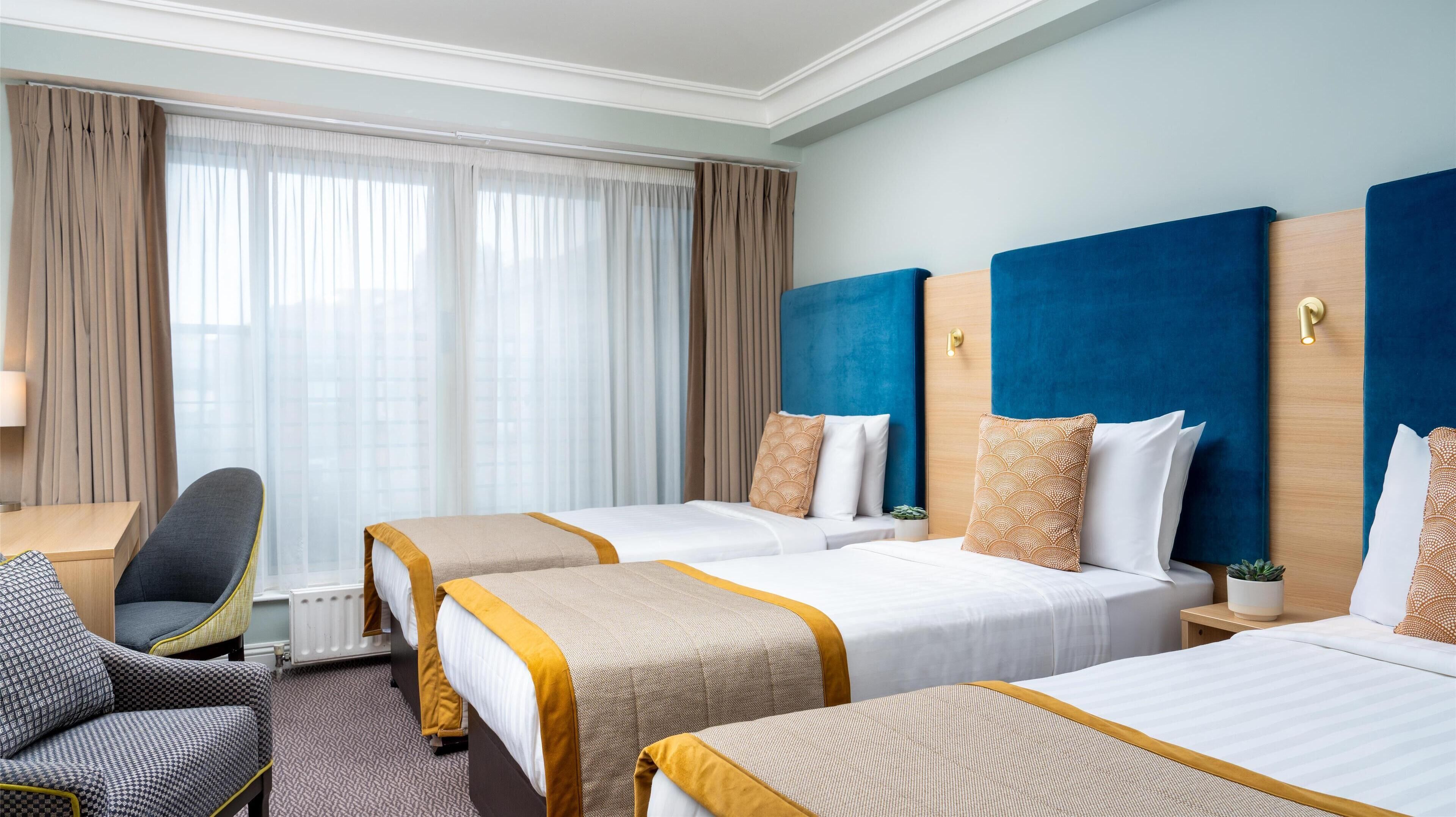 Quarto de hotel com duas camas, uma escrivaninha, uma cadeira e uma janela grande com cortinas.
