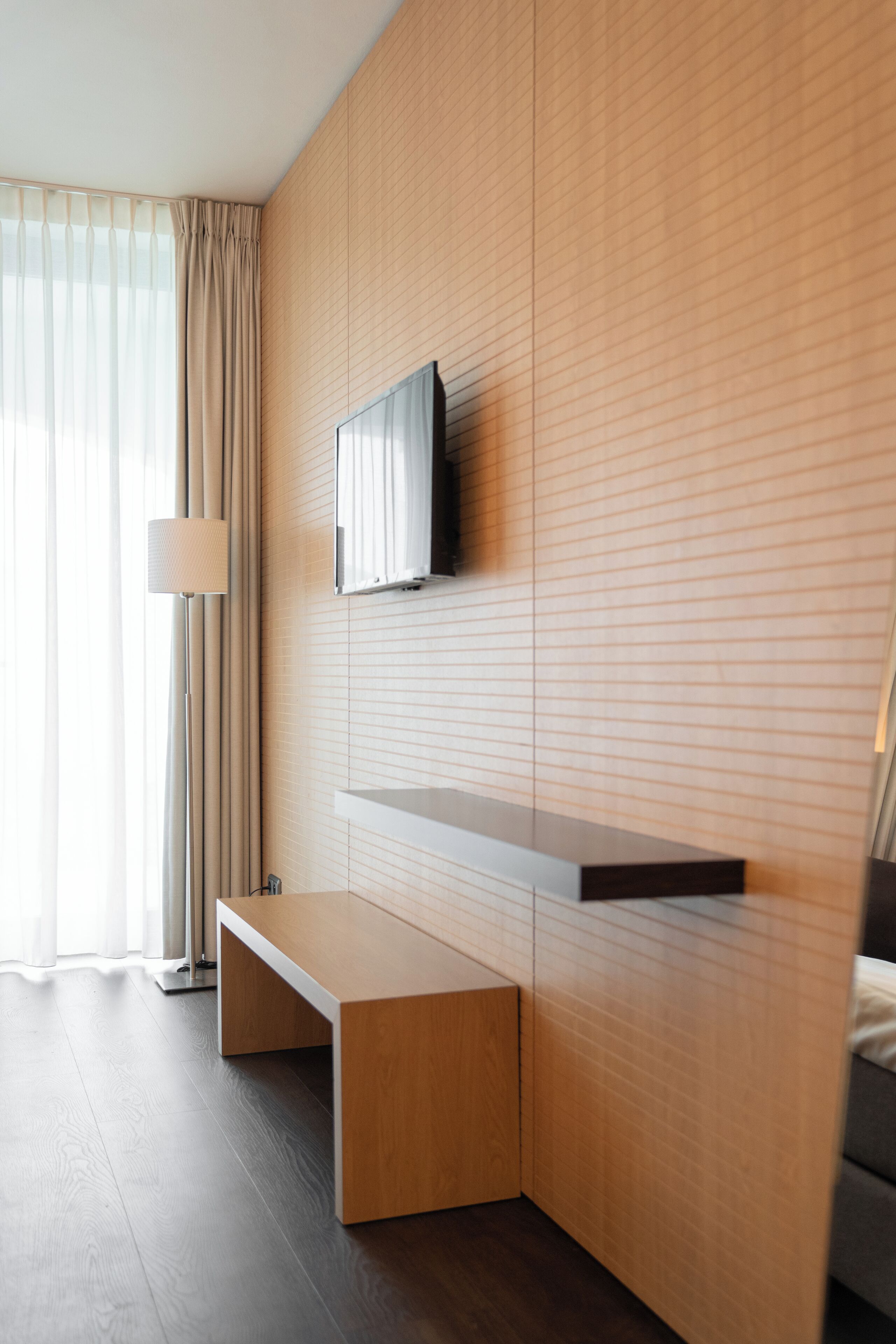 Quarto de hotel moderno com parede de painéis de madeira, TV de tela plana montada, um banco e uma grande janela com cortinas.