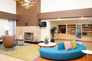 Um lobby moderno com lareira de pedra, televisão de tela plana, área de estar com sofá azul e uma cozinha compacta ao fundo.