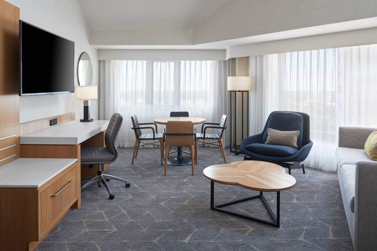 Quarto de hotel com escrivaninha, duas cadeiras, uma mesa redonda, um sofá, televisão e janelas amplas com cortinas translúcidas.