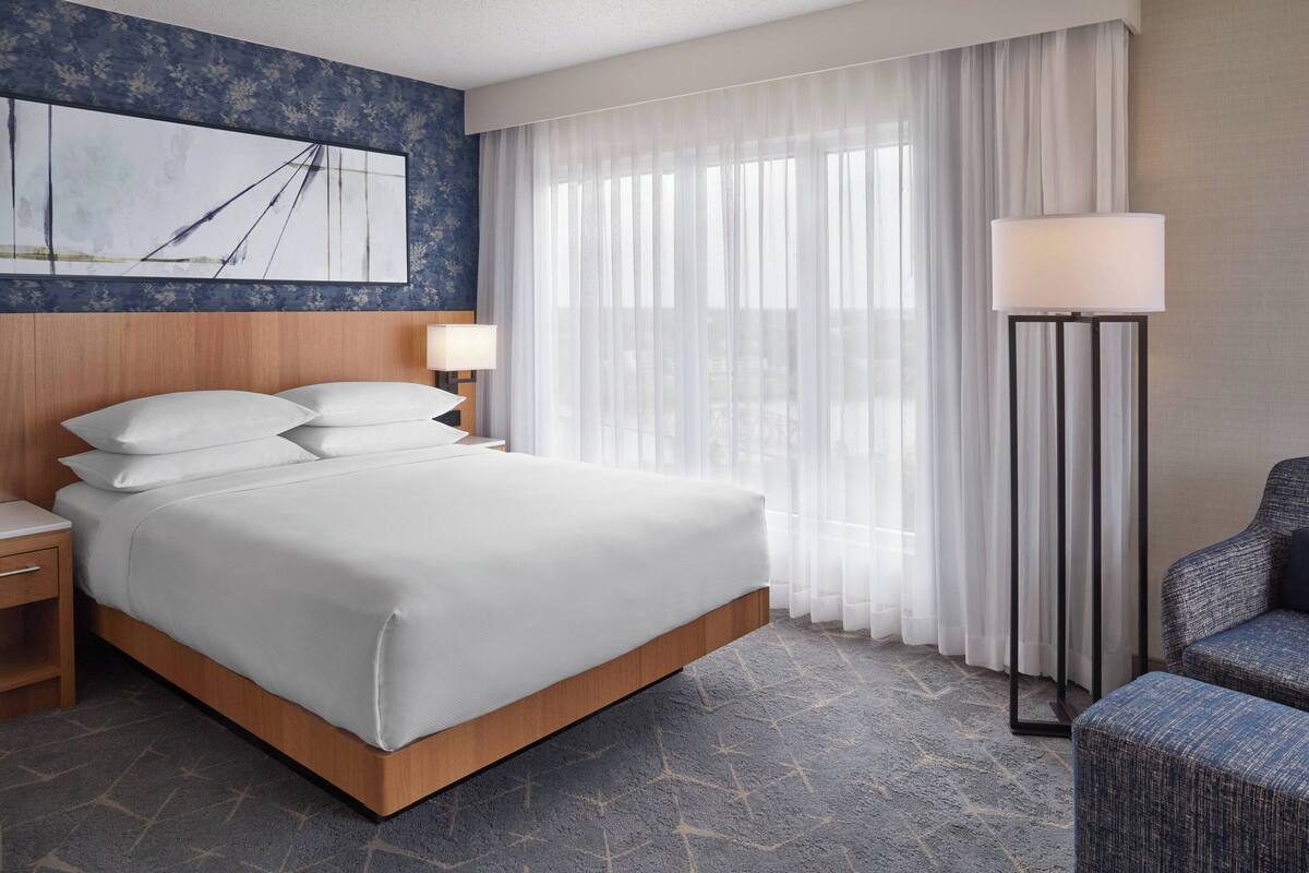 Quarto de hotel com cama, mesa de cabeceira, luminária, cadeira e uma janela grande com cortinas.