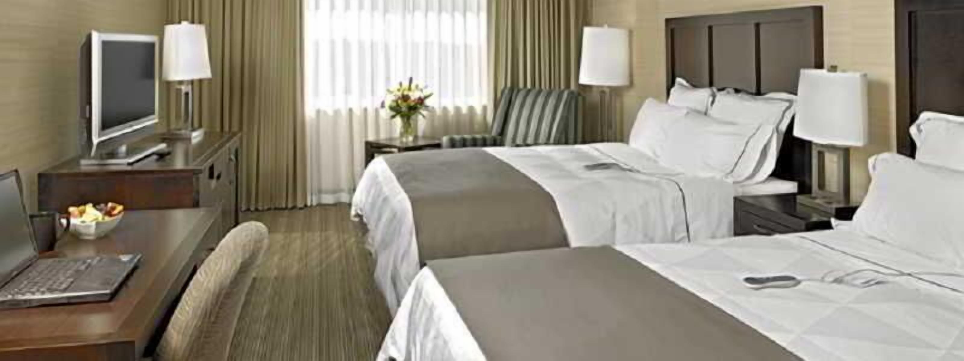 Quarto de hotel com duas camas, uma escrivaninha com um laptop, uma televisão e um vaso com flores.
