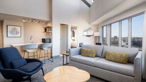 Uma sala de estar moderna com um sofá cinzento, uma poltrona azul e uma mesa de centro de madeira. Há uma área de cozinha com micro-ondas e uma mesa de jantar com duas cadeiras. O quarto possui grandes janelas com vista para a cidade.