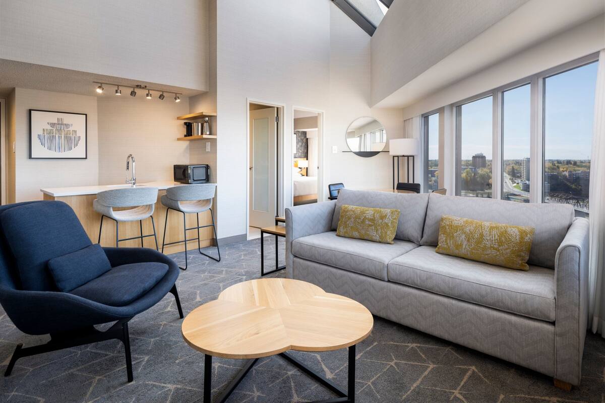 Uma sala de estar moderna com um sofá cinzento, uma poltrona azul e uma mesa de centro de madeira. Há uma área de cozinha com micro-ondas e uma mesa de jantar com duas cadeiras. O quarto possui grandes janelas com vista para a cidade.