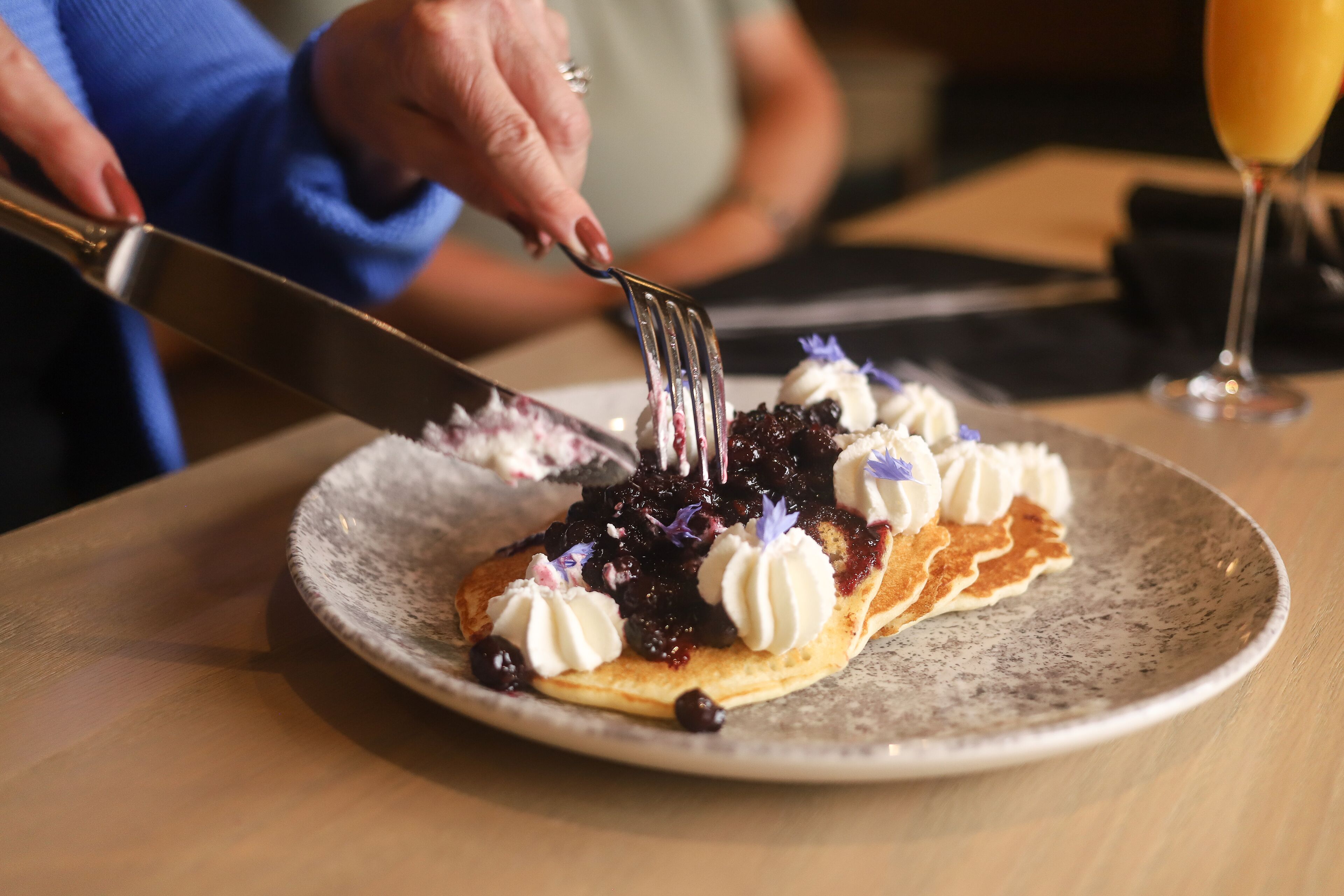 Um prato de sobremesa com uma fatia de bolo coberta com frutas vermelhas e chantilly, um garfo e um copo de suco de laranja sobre uma mesa.