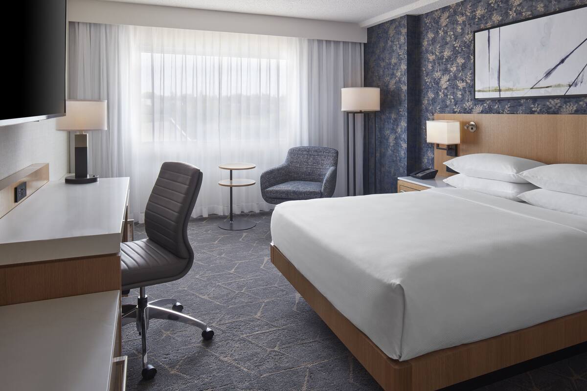 Quarto de hotel com uma cama grande, uma escrivaninha, uma cadeira, duas mesas pequenas e uma televisão.
