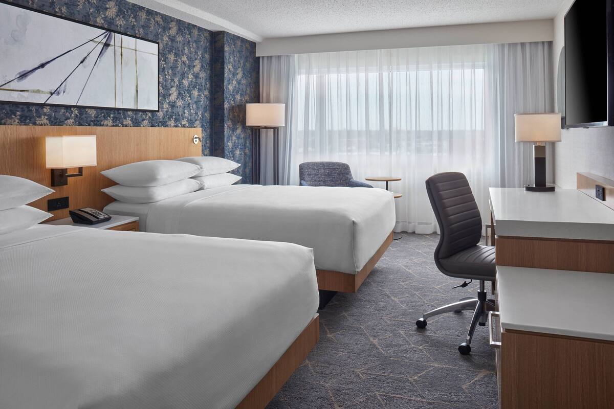 Quarto de hotel com duas camas, uma escrivaninha, uma cadeira, uma televisão e uma janela com cortinas.